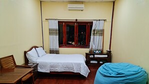 Quarto com cama, mesa de madeira, puff azul e mesa de cabeceira com espelho e pequeno armário.