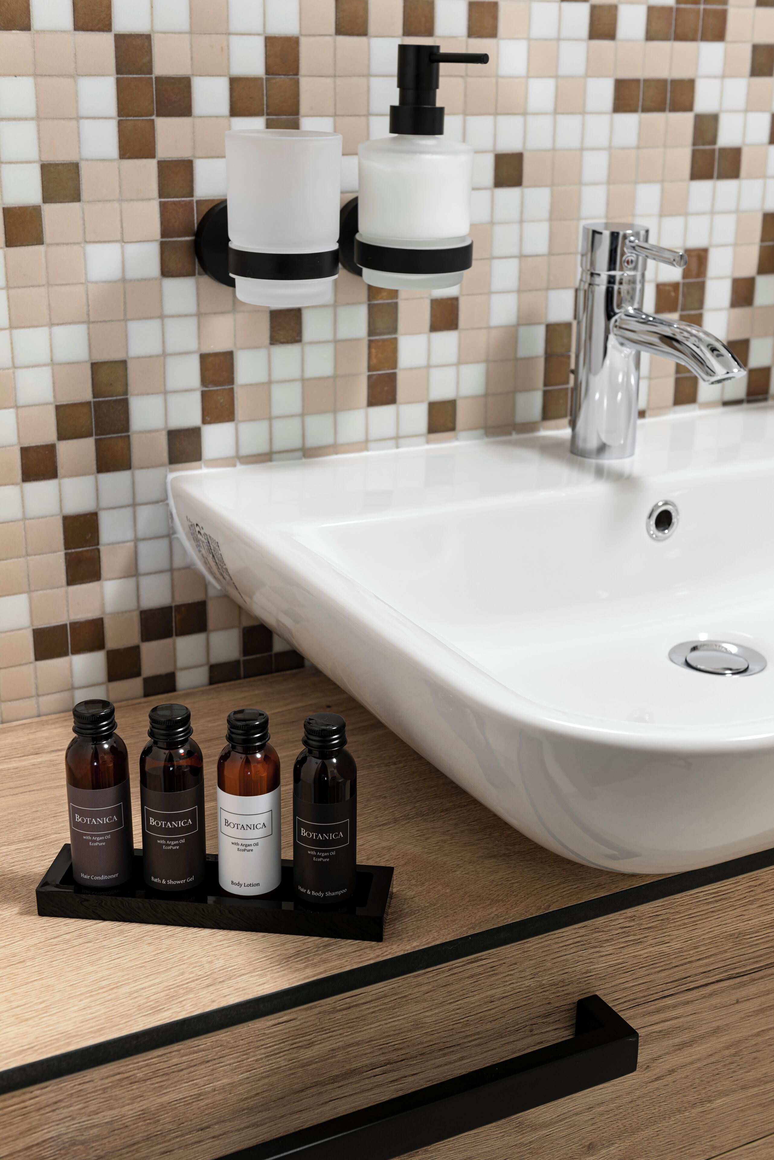 Um banheiro com pia branca, espelho e painel de azulejos. Quatro frascos de produtos de cuidados Botanica estão no balcão.