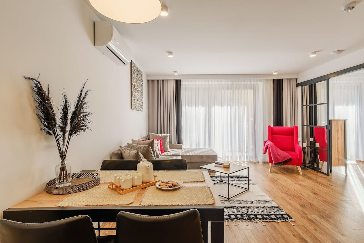Uma sala de estar moderna com área de jantar, apresentando um sofá, uma poltrona vermelha e uma janela grande com cortinas.