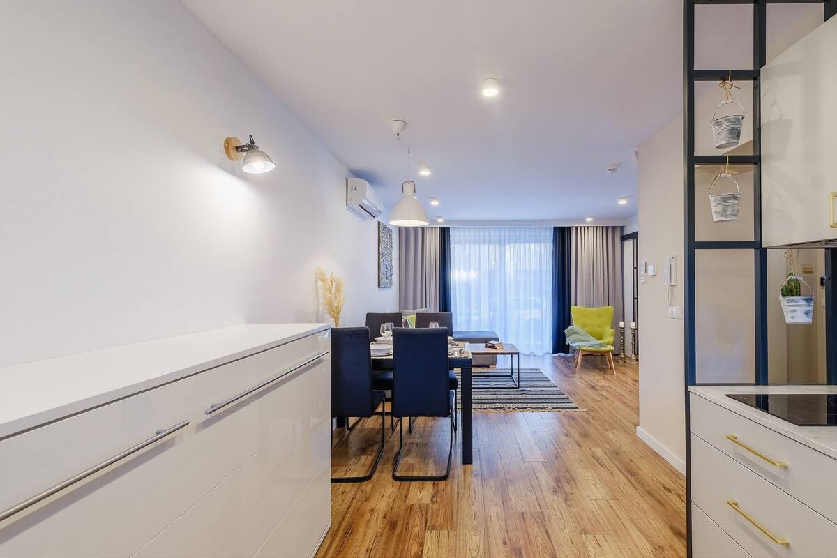 Cozinha moderna com armários brancos, área de refeições com mesa e cadeiras, e uma janela iluminada com cortinas.