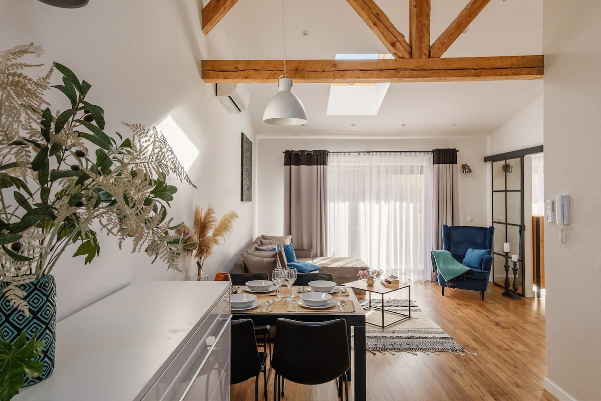Uma sala de estar moderna com área de jantar, vigas de madeira, uma janela grande e uma poltrona azul.
