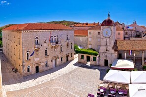 Uma praça histórica com calçamento de pedra, uma imponente torre do relógio e edifícios tradicionais com varandas e bandeiras.