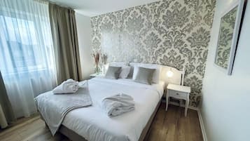 Cama bem arrumada com travesseiros e manta, mesa de cabeceira com abajur e papel de parede estampado na parede.