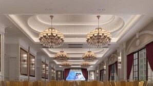 Uma sala de banquetes com fileiras de mesas e cadeiras, grandes lustres e um palco com uma tela.