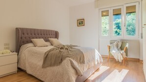 Quarto com cama grande, mesa de cabeceira, cadeira e janela com vista para área verde.