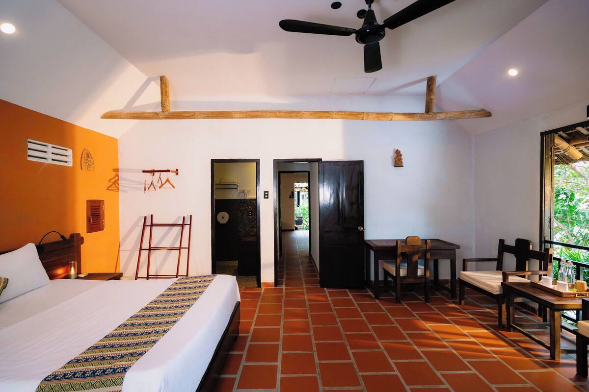 Quarto de hotel com cama, escrivaninha e cadeira. Há um ventilador de teto e uma janela com vista para o verde.