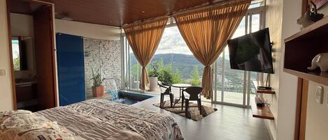 Quarto com cama grande, televisão, mesinha com cadeiras, planta em vaso e vista para o exterior.