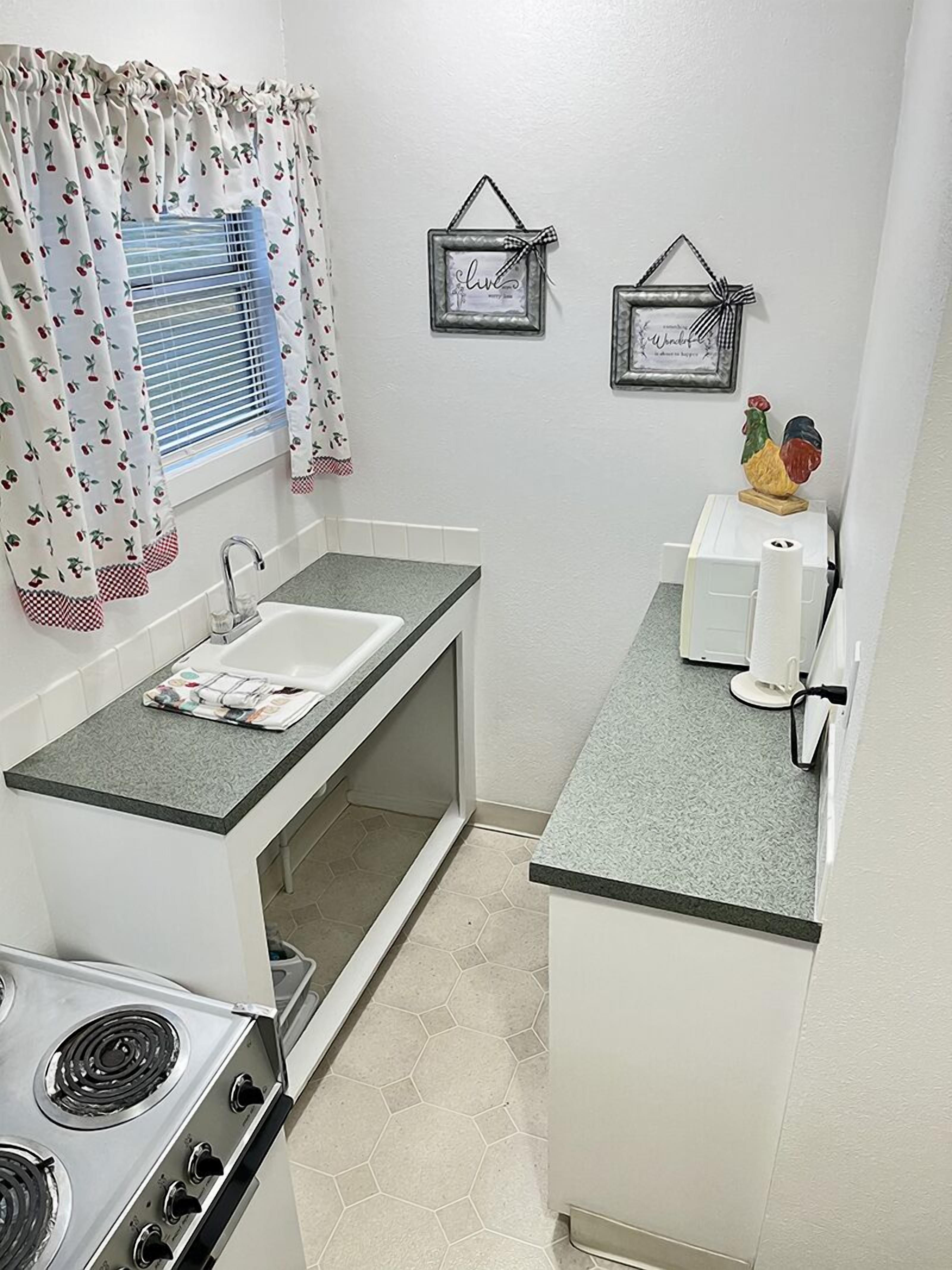 Cozinha compacta com fogão, pia e torradeira. Objetos decorativos na parede e uma janela com cortinas.