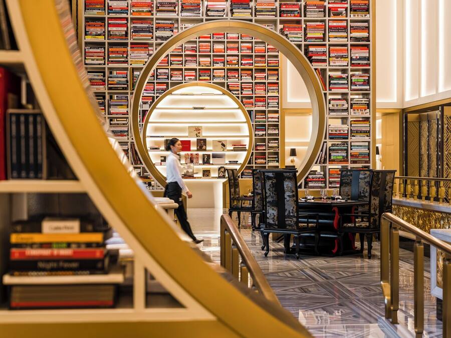 Uma biblioteca com estantes, um elemento decorativo circular e uma pessoa caminhando.