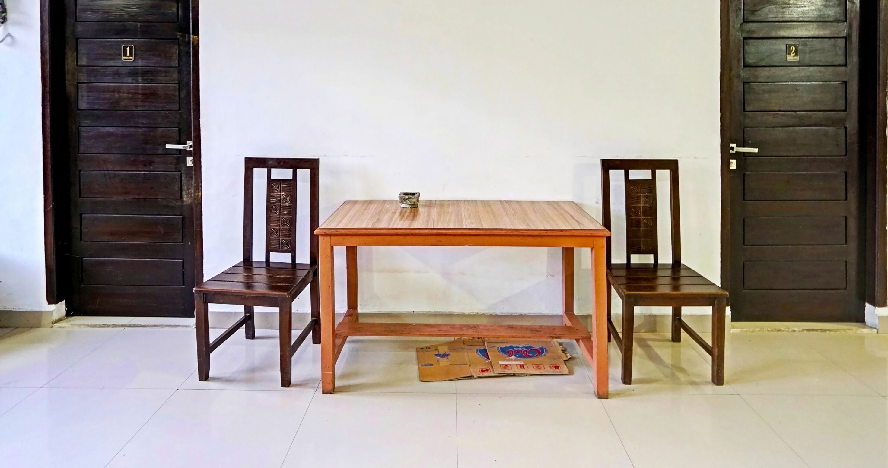 Quarto com mesa de madeira e duas cadeiras, chão de azulejos e duas portas de madeira escura com números.