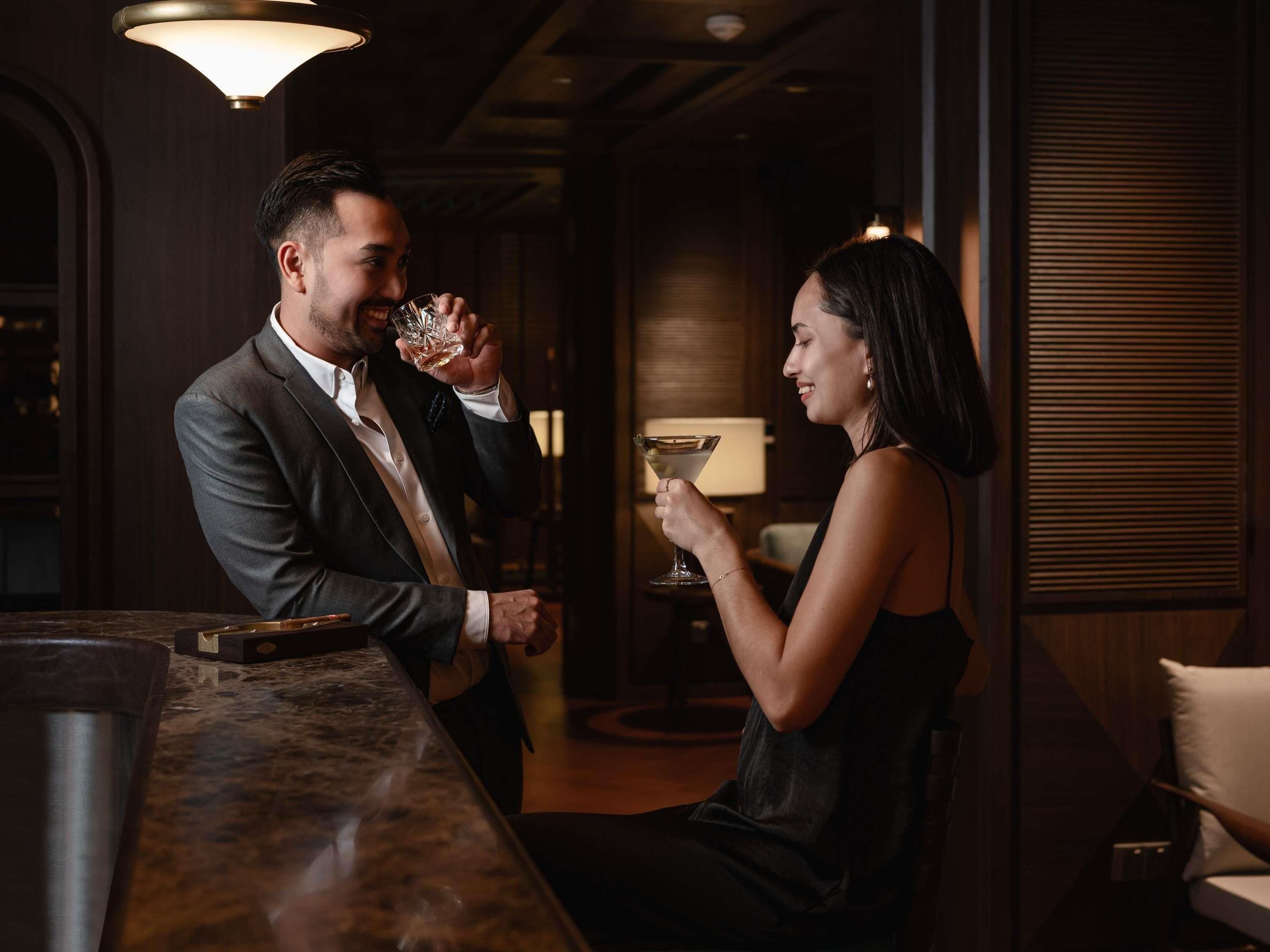 Um homem e uma mulher num bar com bancada de mármore, uma lâmpada e uma parede de madeira ao fundo.