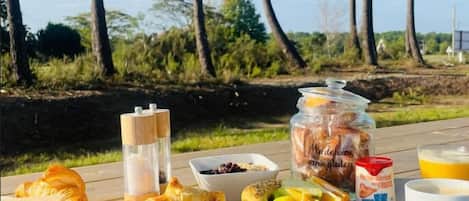 Café da manhã ao ar livre com diversos alimentos e bebidas sobre uma mesa, cercada por árvores.