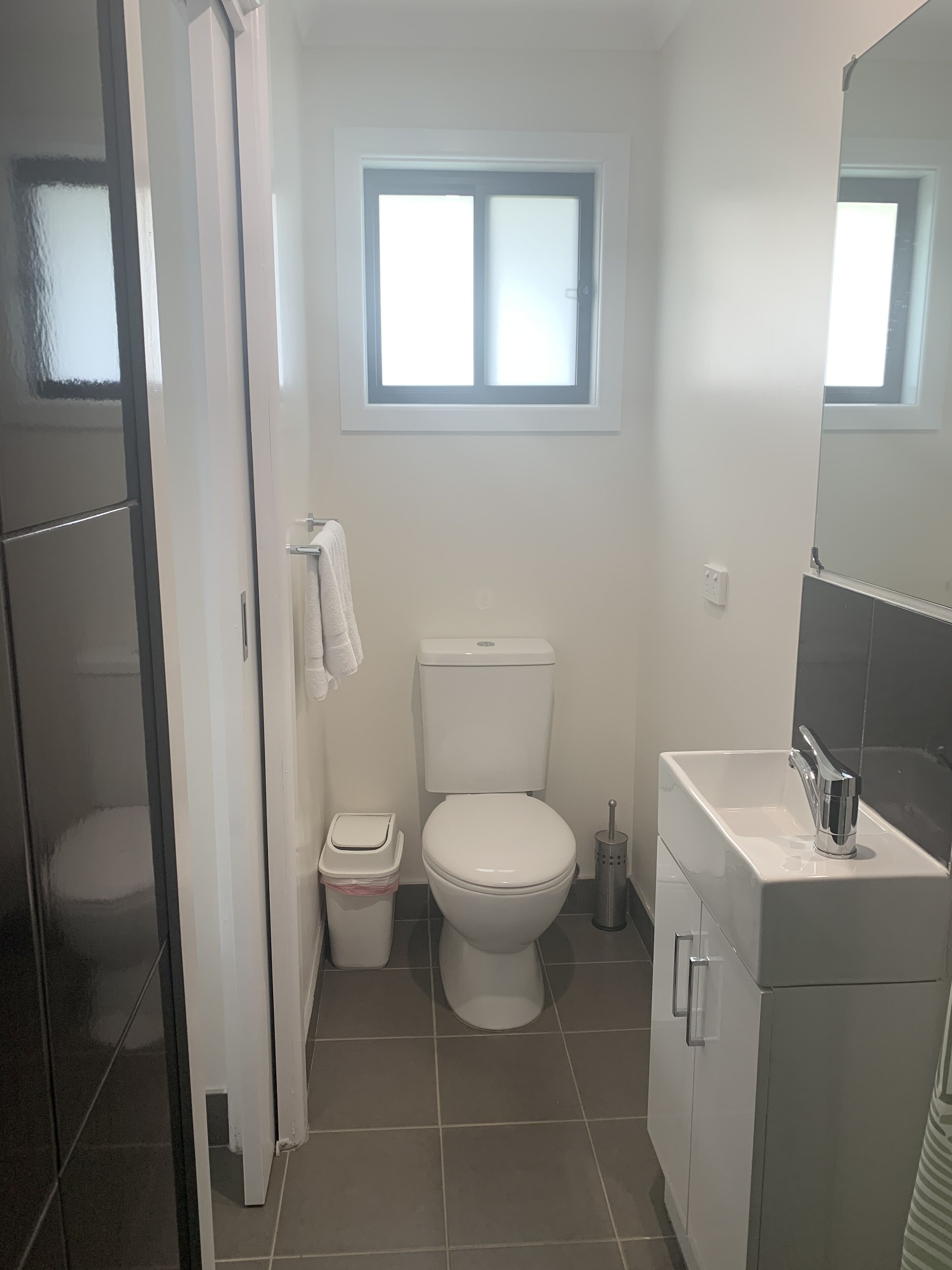 Uma casa de banho pequena com sanita, uma janela, lavatório e um cesto de lixo.