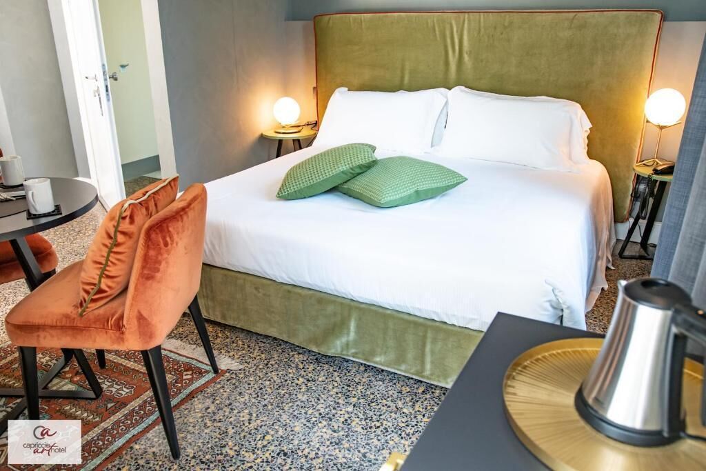 Quarto de hotel com uma cama, duas luminárias de cabeceira, uma mesa de centro, uma cadeira e uma chaleira.