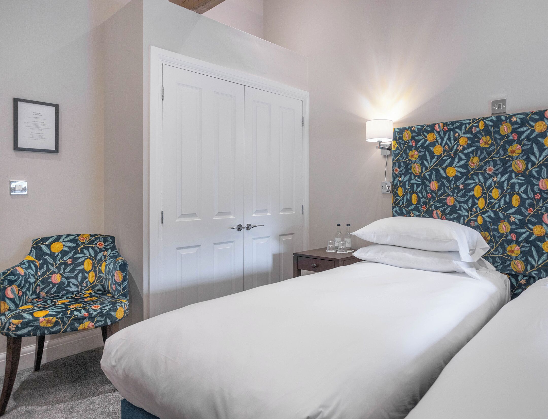 Quarto de hotel com uma cama, uma cadeira, um guarda-roupa e uma mesa de cabeceira com abajur.