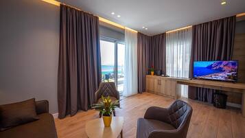 Uma sala de estar moderna com sofá, poltrona e uma mesinha com uma planta. Uma televisão de tela plana está fixada na parede, e visível está uma varanda com cortinas.