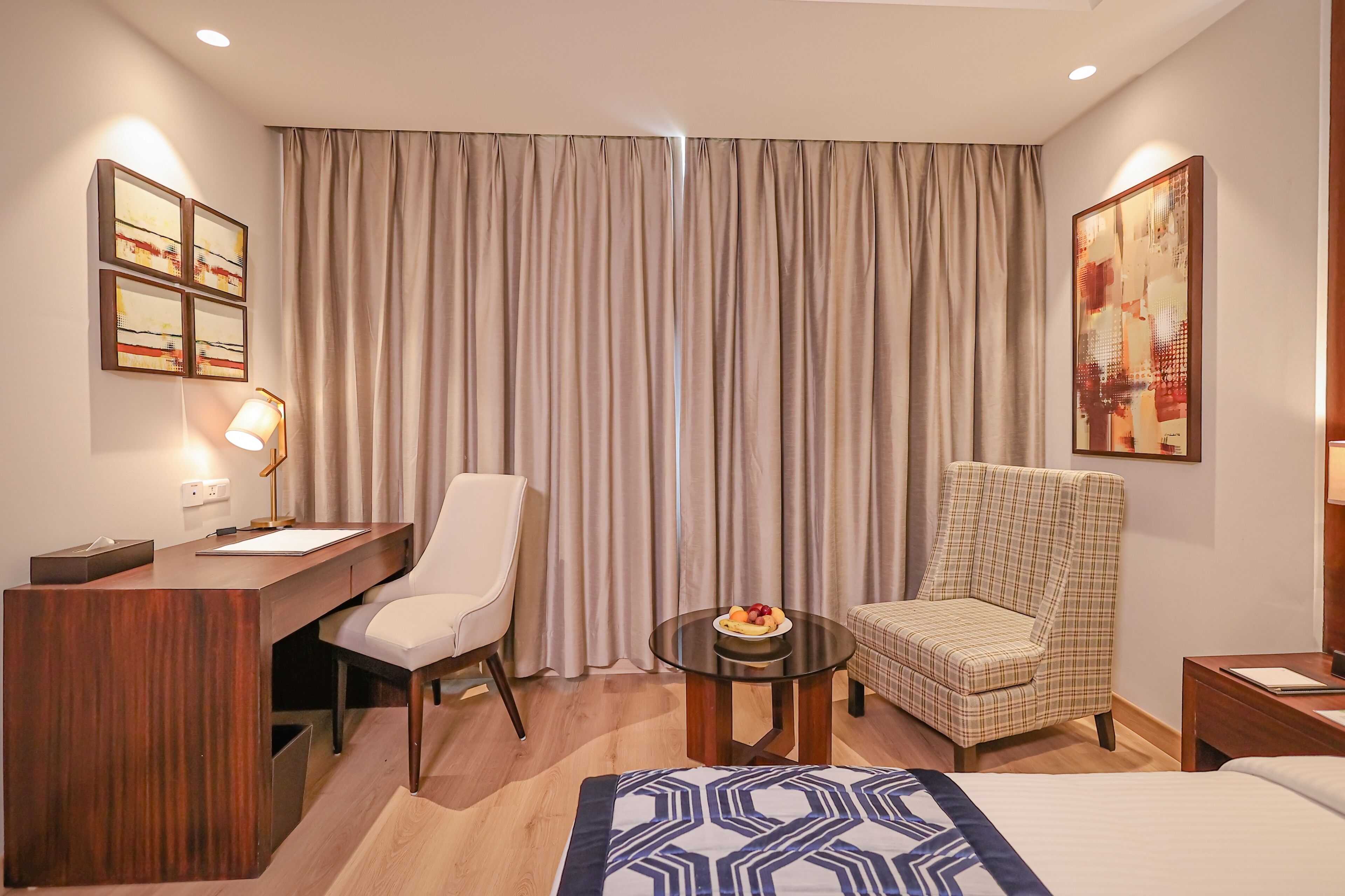Quarto de hotel com secretária de madeira, uma cadeira, uma cama com colcha estampada, uma pequena mesa redonda com frutas e uma grande janela com cortinas.