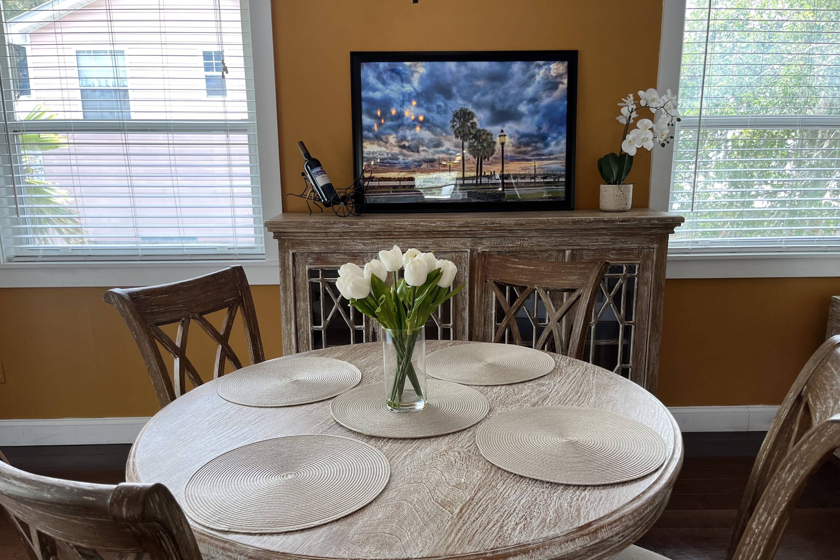 Mesa de jantar com um vaso de flores brancas, uma televisão sobre um móvel de madeira e uma janela com persianas.