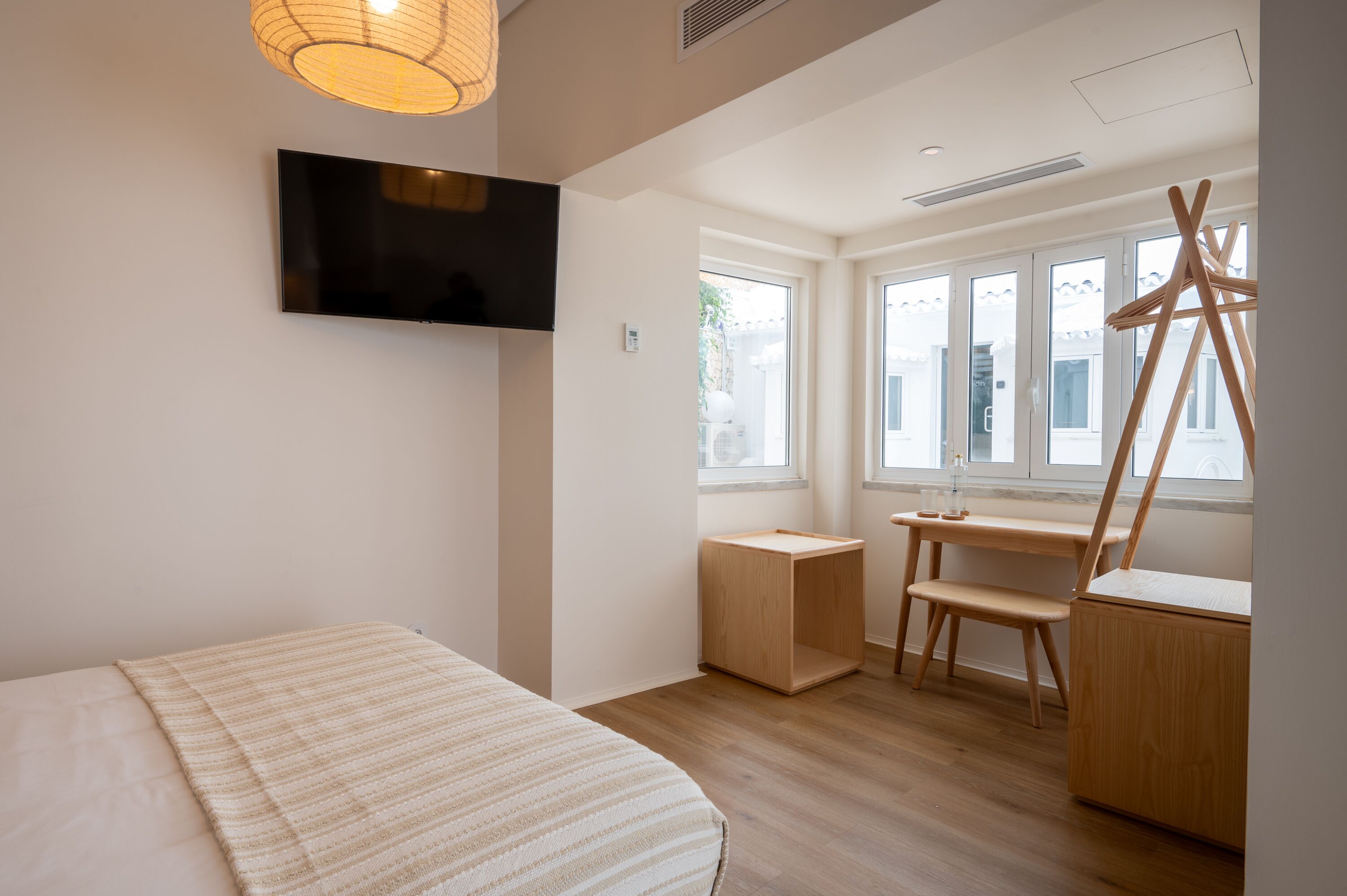 Quarto moderno com cama, televisão na parede, escrivaninha com cadeira e janelas amplas.