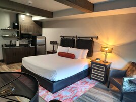 Quarto de hotel moderno com cama grande, escrivaninha pequena, uma cadeira, área de cozinha e banheiro.