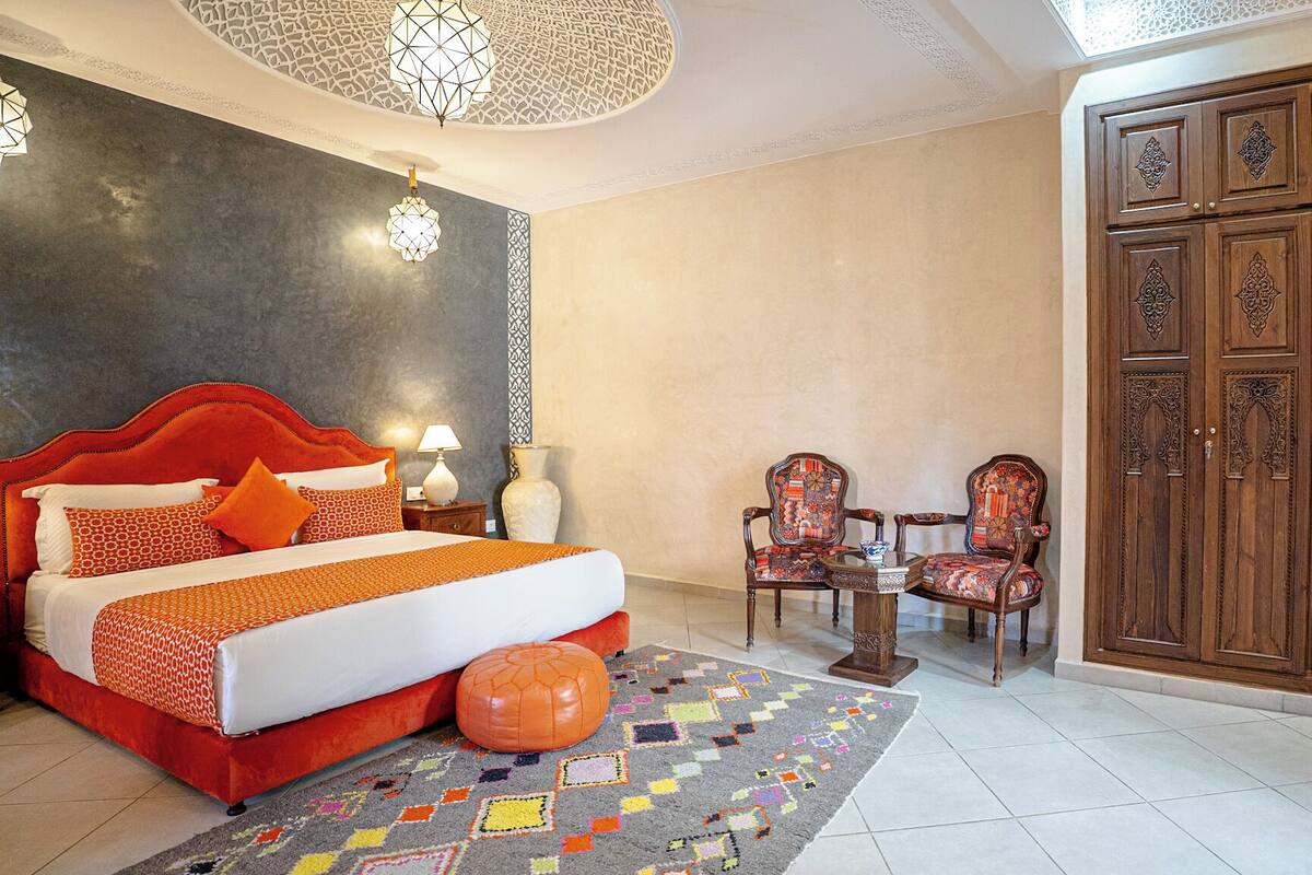 Quarto de hotel com uma cama grande, duas cadeiras, um guarda-roupa e um tapete estampado.