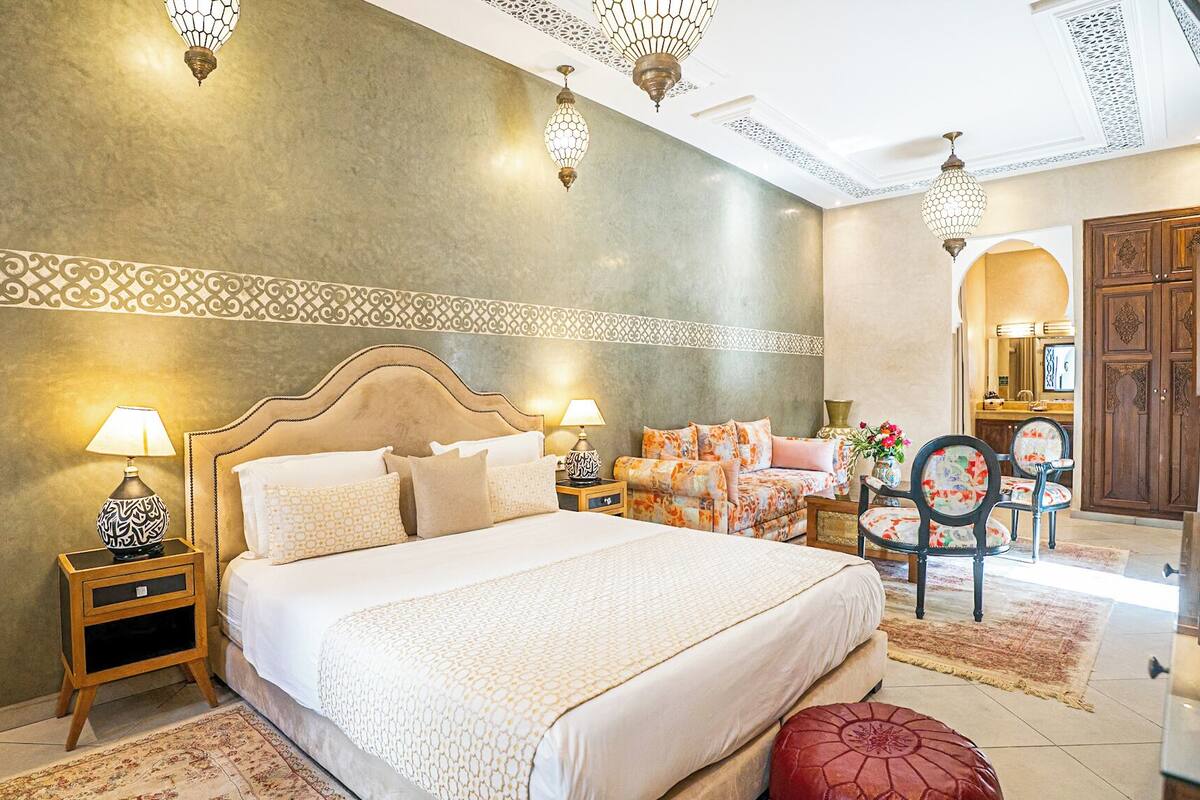 Quarto com uma cama grande, duas mesas de cabeceira com luminárias, uma cadeira, uma mesinha com um vaso e um guarda-roupa.