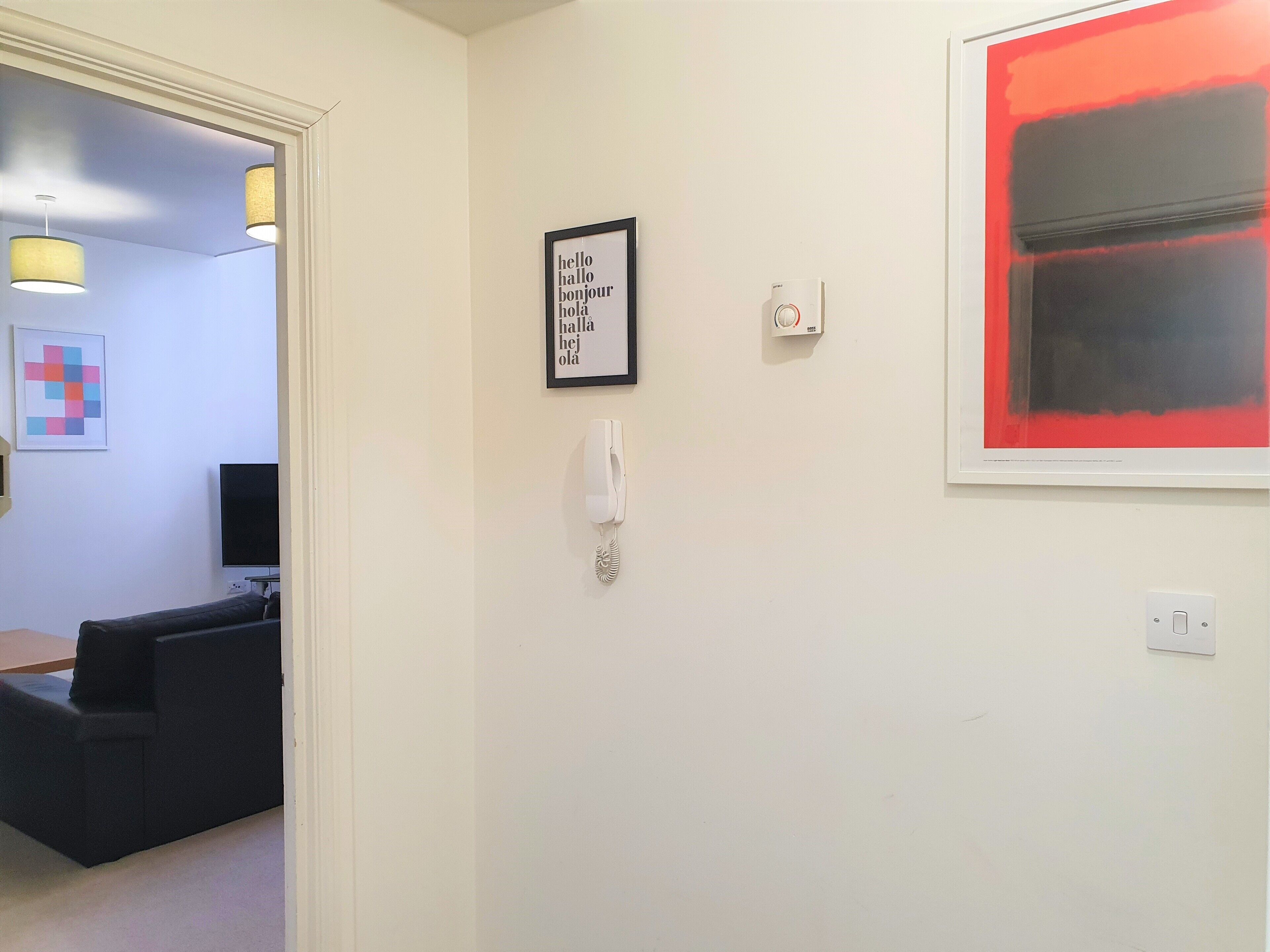 Quarto com sofá preto, placa de saudação multilíngue emoldurada, telefone branco e obra de arte vermelha e preta na parede.