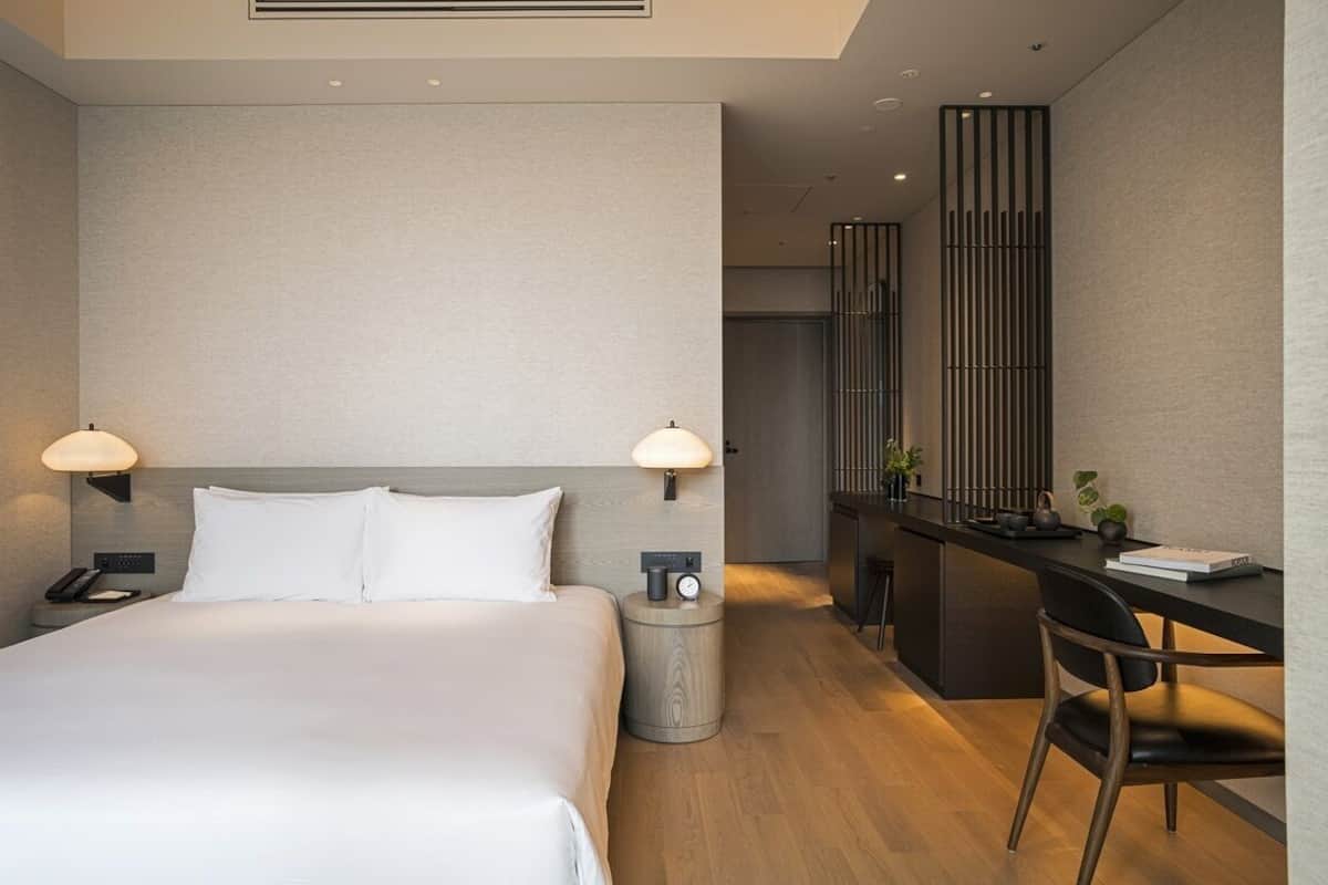 Quarto de hotel moderno com uma cama grande, abajures, uma mesa de escritório com cadeira e uma pequena mesinha de apoio.
