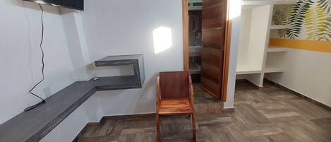 Quarto com piso de madeira, televisão fixada na parede, uma cadeira de madeira e uma porta entreaberta que dá para outro espaço.