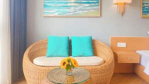 Uma cadeira de vime com uma mesa de vidro e um vaso de girassóis, um quadro de um pôr do sol sobre o oceano e uma mesa de cabeceira com uma luminária.