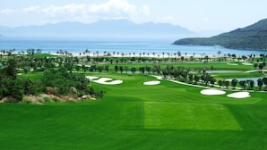 Um campo de golfe com bunkers de areia, um corpo de água e montanhas ao longe.