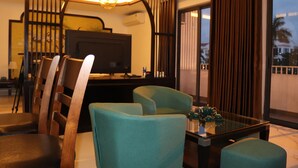 Quarto de hotel moderno com área de jantar, mesa de vidro, cadeiras azul-turquesa e uma televisão.