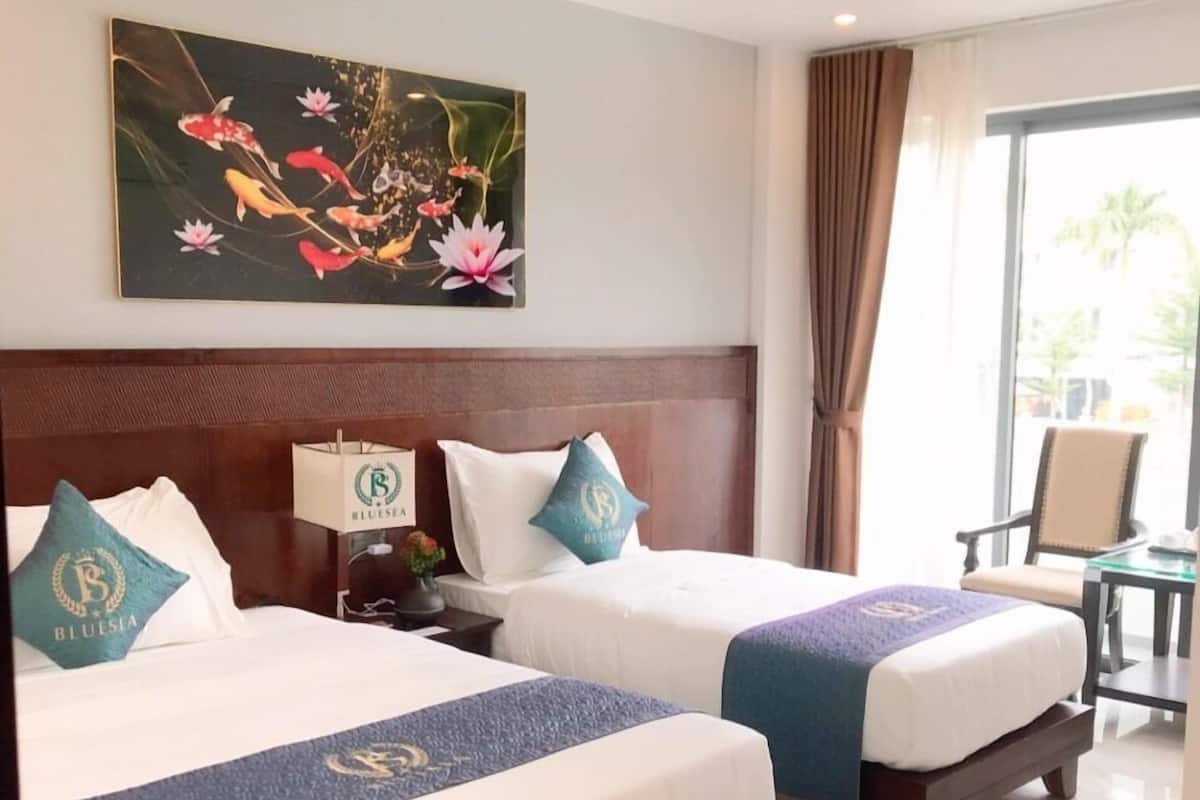 Quarto de hotel com duas camas, mesa de cabeceira com abajur, quadro na parede e vista para o exterior.