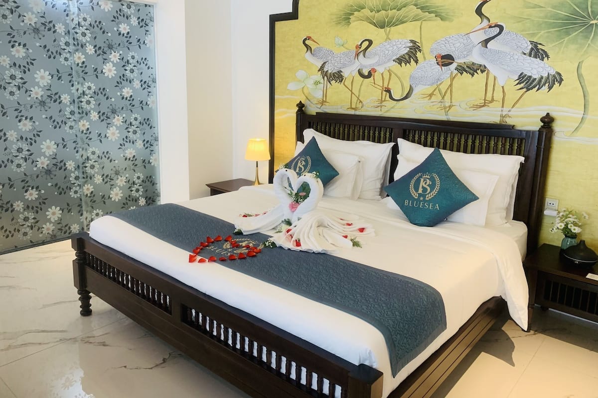 Quarto de hotel com cama, papel de parede floral, cabeceira com pintura de grous e mesa de cabeceira com abajur.