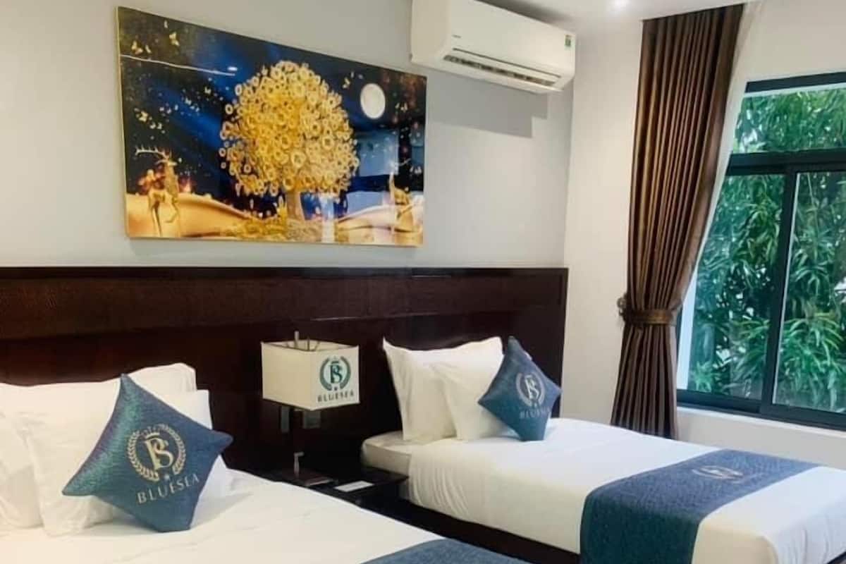 Quarto de hotel com duas camas, mesa de cabeceira, um quadro, ar condicionado e janela com cortinas.
