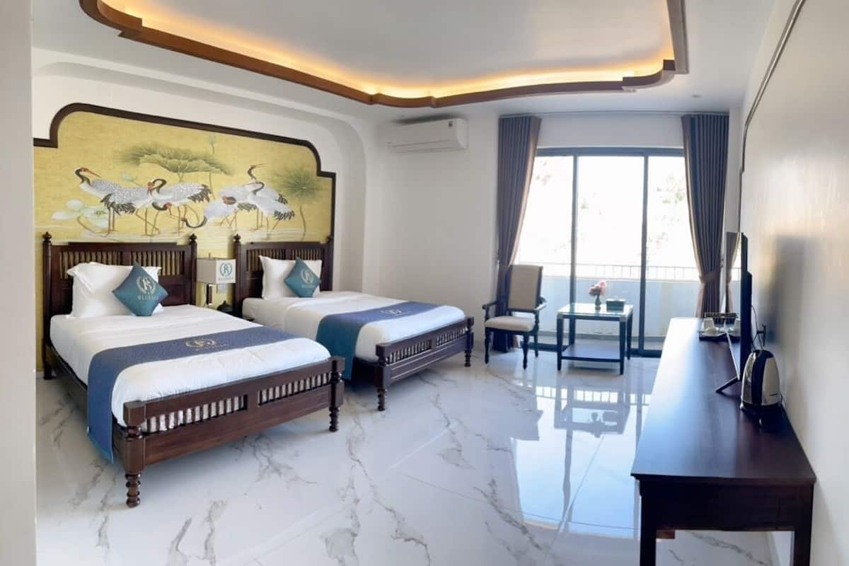 Quarto de hotel com duas camas, uma escrivaninha, uma cadeira, varanda com vista e um quadro na parede.