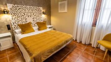 Uma cama bem arrumada com cabeceira estampada, duas mesas de cabeceira, uma cadeira e uma janela com cortinas.