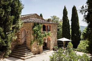 Um edifício de pedra com telhado em tijolos, escadaria que dá acesso à entrada e área de jardim com mesa e cadeiras.
