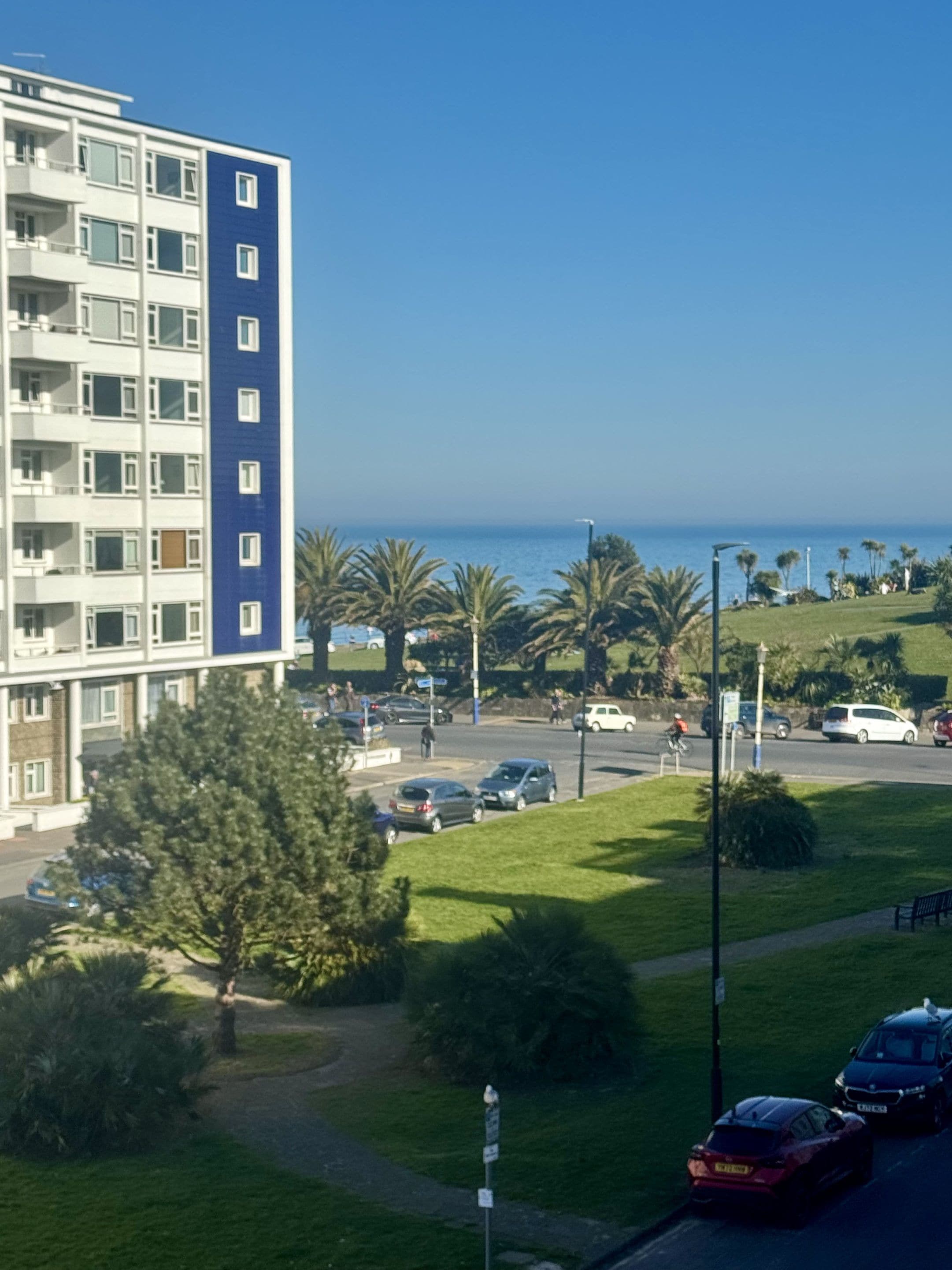 Um edifício de vários andares com fachada azul e branca, situado perto de uma estrada com carros estacionados e palmeiras.