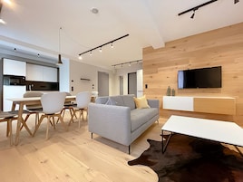 Uma sala de estar moderna com sofá cinzento, soalho de madeira, área de refeições com cadeiras, cozinha ao fundo e uma TV na parede.