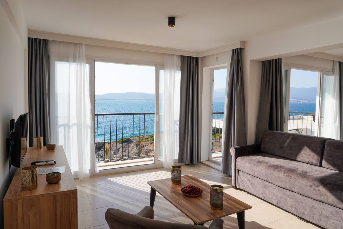 Uma sala de estar ampla com um sofá, uma mesa de centro de madeira e uma televisão. Janelas grandes oferecem vista para o mar e uma varanda.
