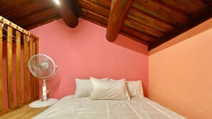 Quarto com cama, travesseiros, ventoinha, vigas de madeira e parede rosa.