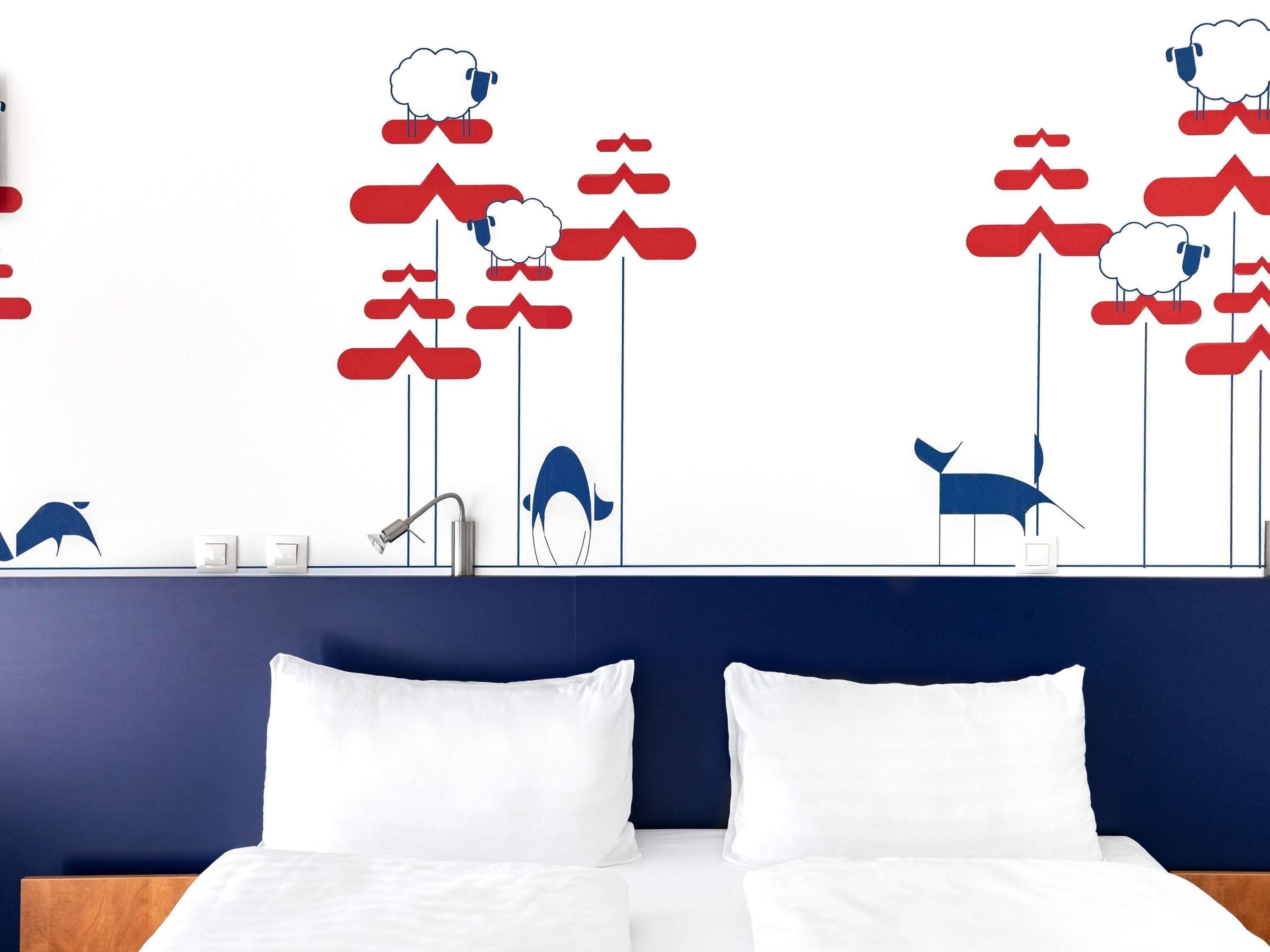 Quarto de hotel com cama, duas almofadas, mesa de cabeceira, espelho e arte na parede com animais estilizados e formas abstratas.