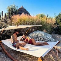 Um casal relaxando em uma rede em um jardim com uma estrutura de telhado de palha ao fundo.