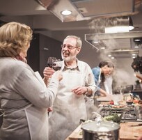 Uma pessoa de avental branco está em uma cozinha com fogão e panelas, cercada por pessoas e uma mesa de jantar com comida e copos de vinho.