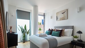 Quarto moderno com uma cama grande, uma mesinha de cabeceira, um vaso de planta e vista para área externa por uma porta deslizante.