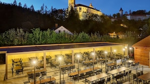 Uma área de refeições ao ar livre com mesas e cadeiras, uma seção coberta e um castelo ao fundo.