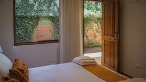 Quarto de hotel com uma cama, janela com vista para uma parede de tijolos e vegetação, e uma porta de madeira.