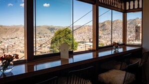 Quarto com uma grande janela que dá para uma vista urbana e montanhas. Há uma mesa de madeira com cadeiras e um vaso de flores sobre ela.