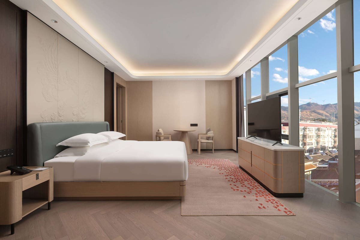 Quarto de hotel moderno com uma cama grande, uma escrivaninha, uma televisão e vista da cidade através de amplas janelas.